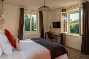 Quarto com cama, escrivaninha, cadeira, televisão e janelas amplas com cortinas.