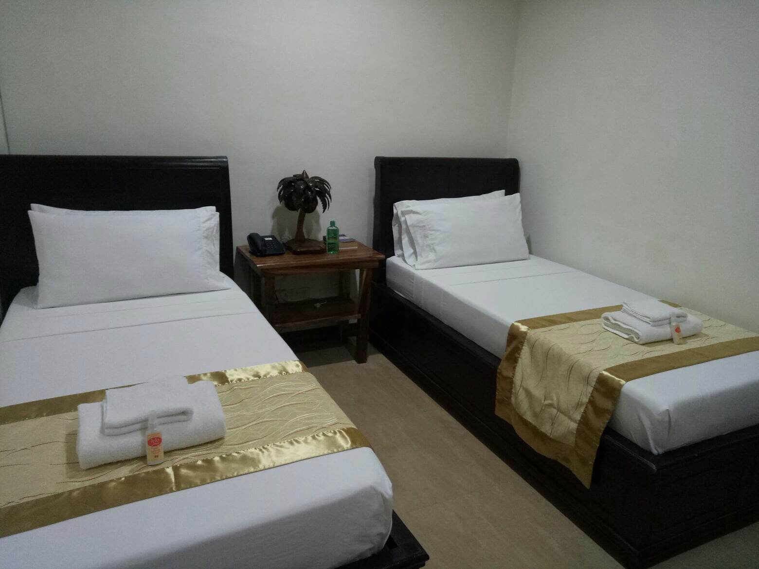 Quarto de hotel com duas camas de solteiro, cada uma com roupa de cama branca e detalhes dourados. Há uma mesa de cabeceira de madeira entre as camas, com um abajur e uma garrafa de água.