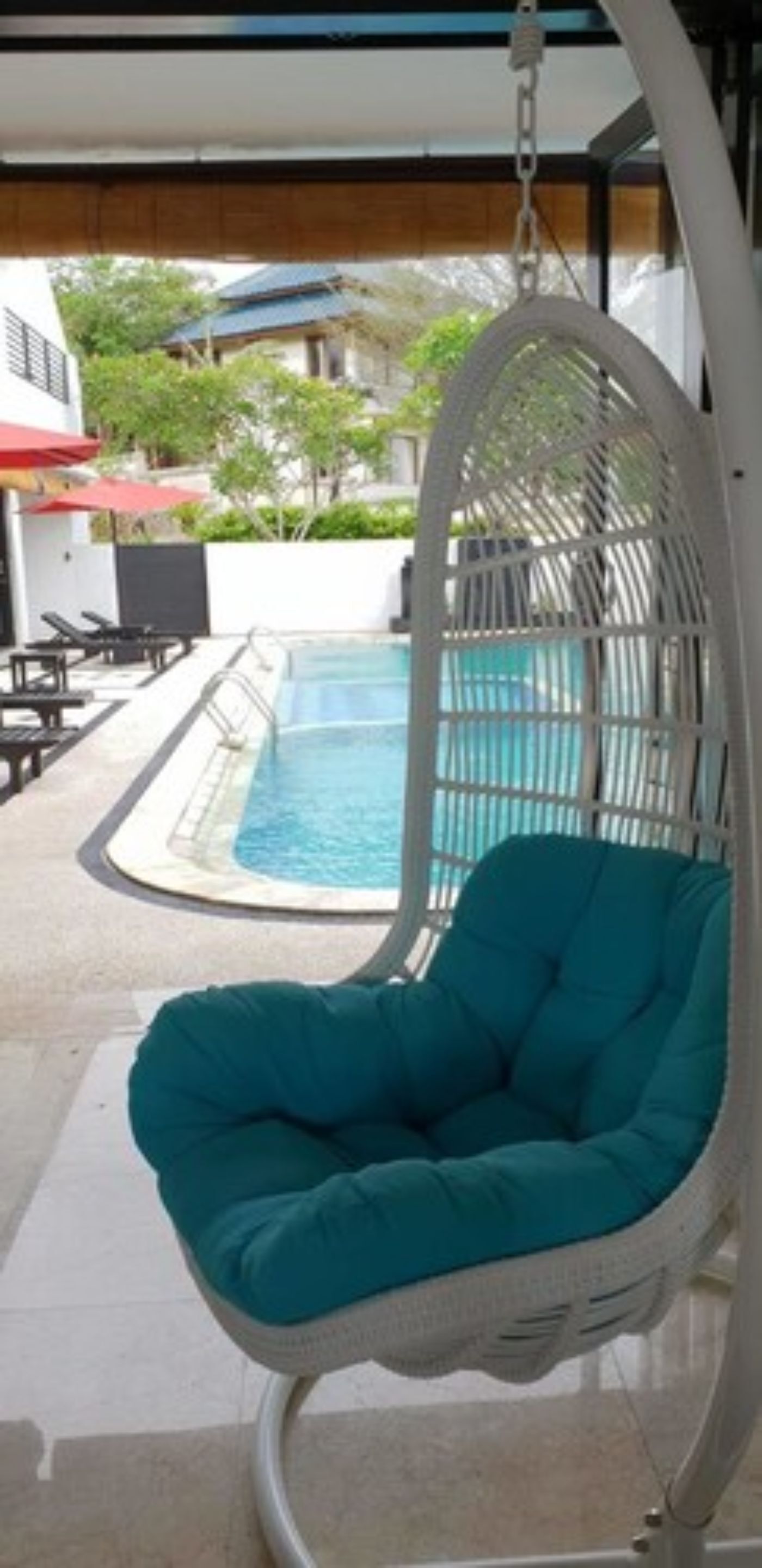 Uma cadeira suspensa com almofada azul-turquesa perto de uma piscina, com uma piscina e um edifício ao fundo.