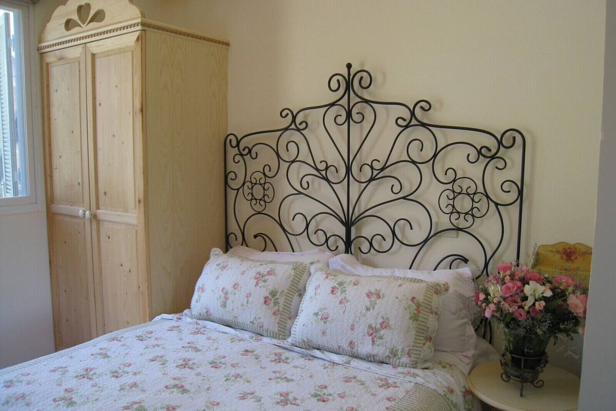 Quarto com cama, roupa de cama floral, cabeceira decorativa, guarda-roupa de madeira e uma pequena mesa com um vaso de flores.