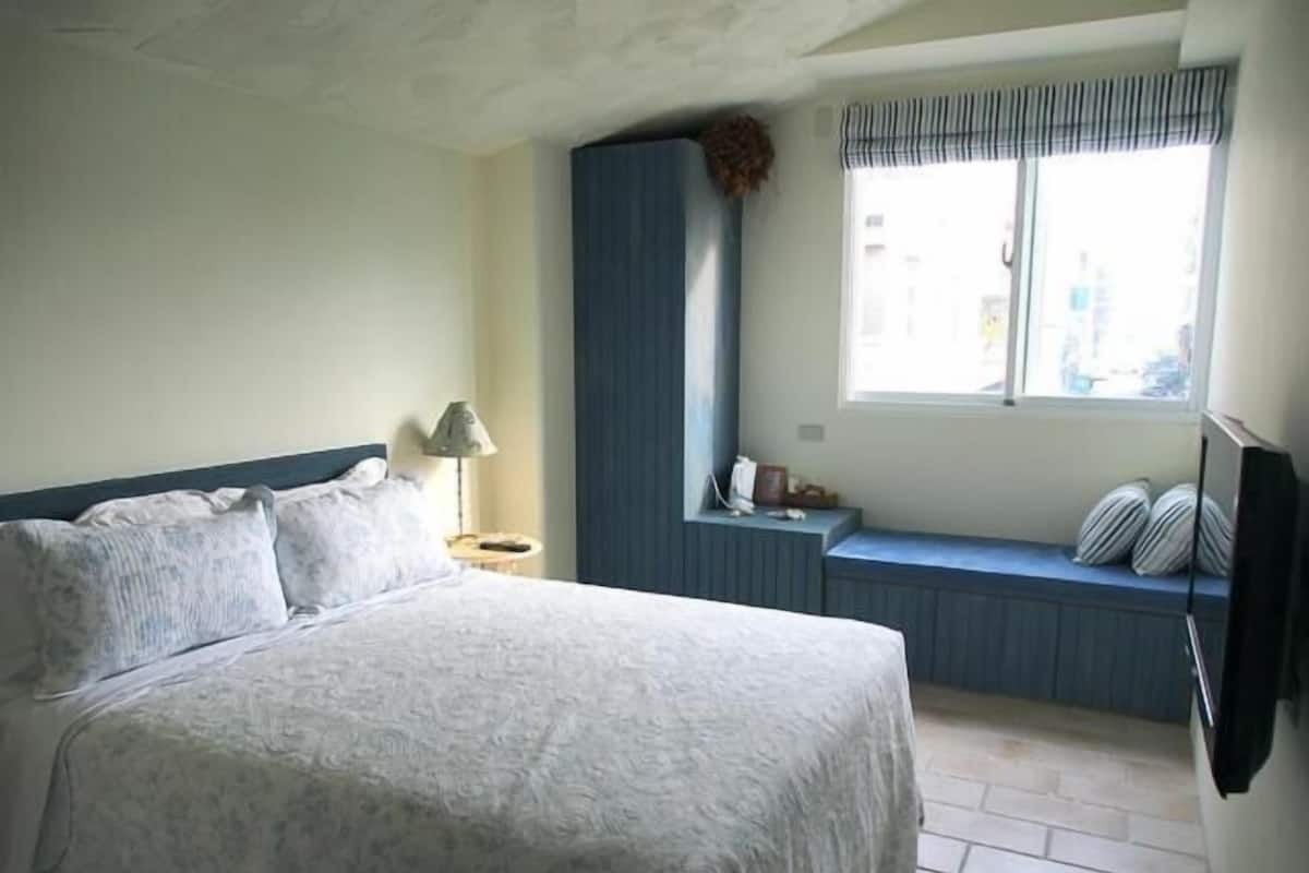 Quarto com uma cama grande, mesa de cabeceira com abajur, janela com vista e um banco embutido com almofadas.