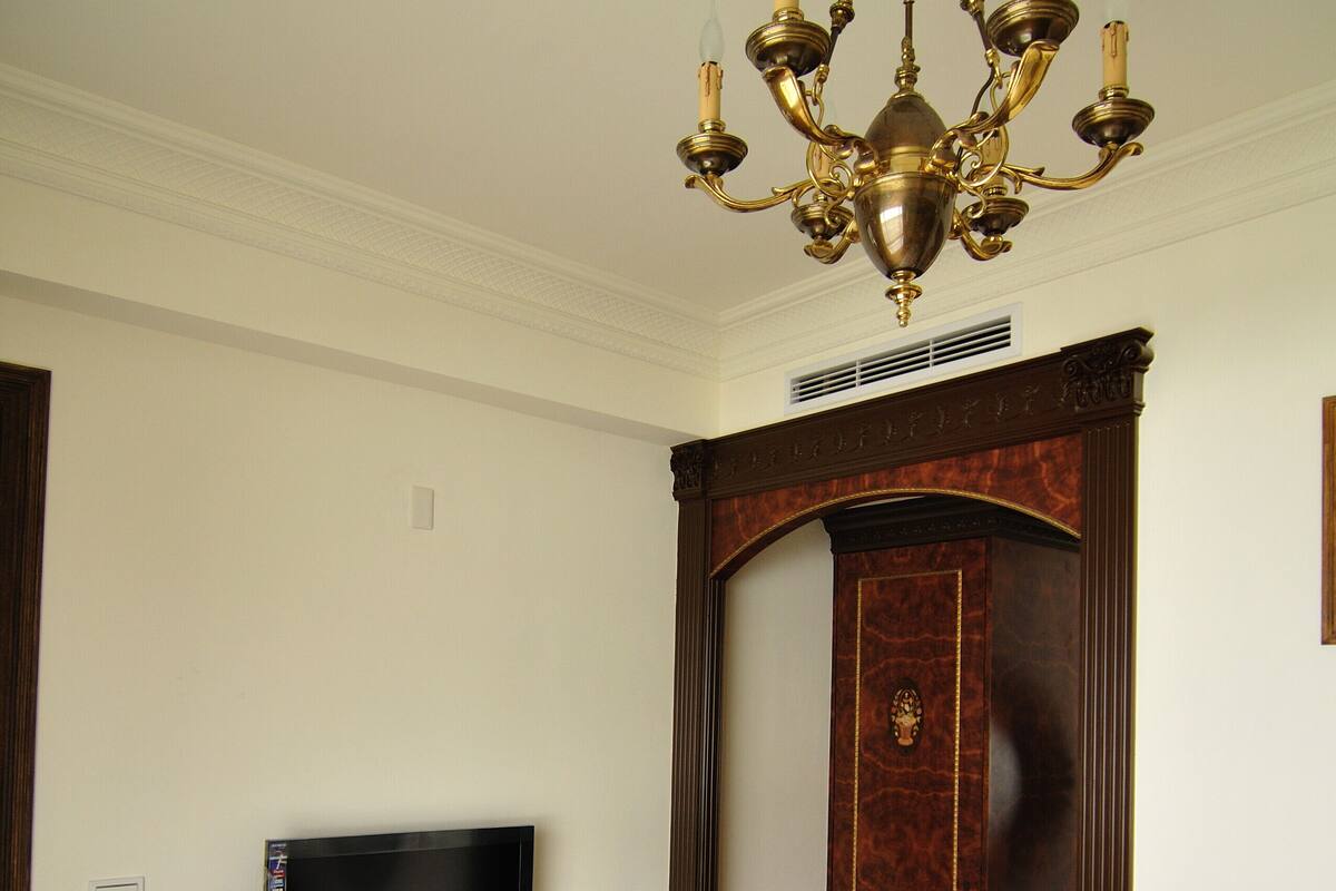 Quarto com cômoda de madeira, televisão, lustre e porta com arco decorativo.
