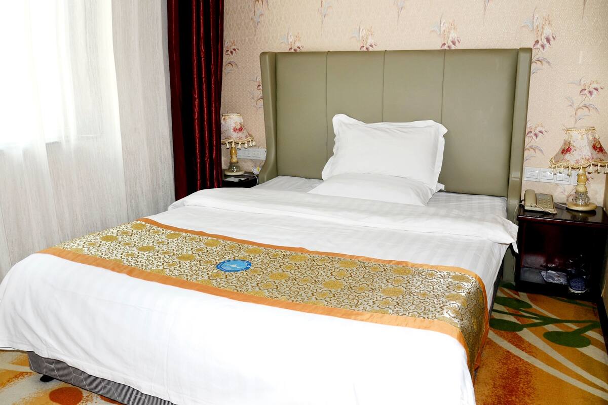 Cama bem arrumada com colcha estampada, mesa de cabeceira com abajur e telefone.