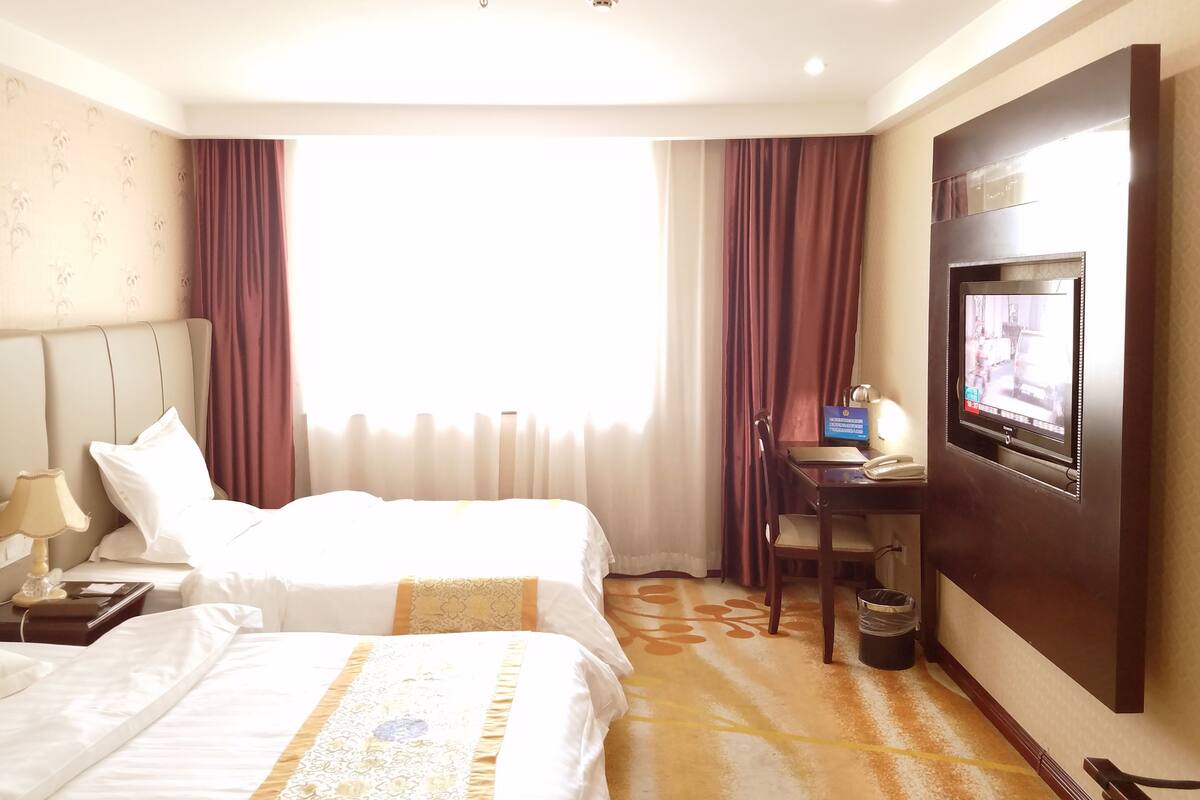 Quarto de hotel com duas camas, uma escrivaninha, televisão e vista exterior através de grandes janelas.