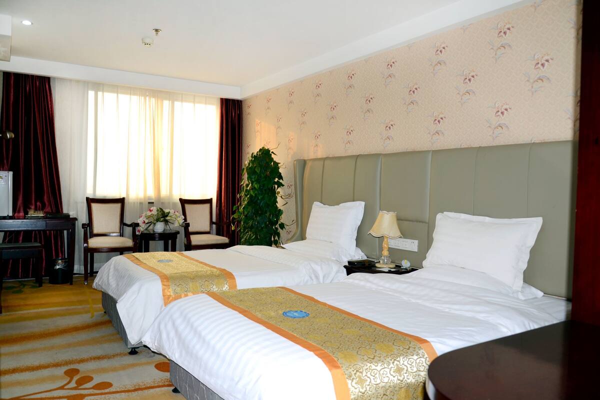 Quarto de hotel com duas camas, uma escrivaninha, cadeiras, uma planta e uma janela com cortinas.