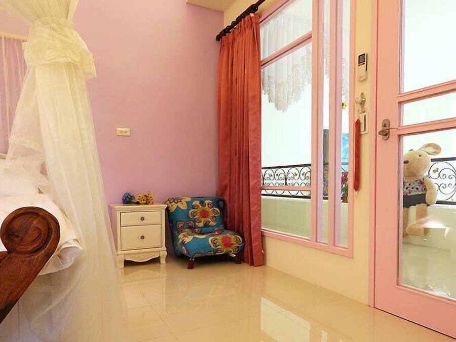 Um quarto com cama de dossel, mesa de cabeceira, cadeira colorida e uma janela grande com cortinas.