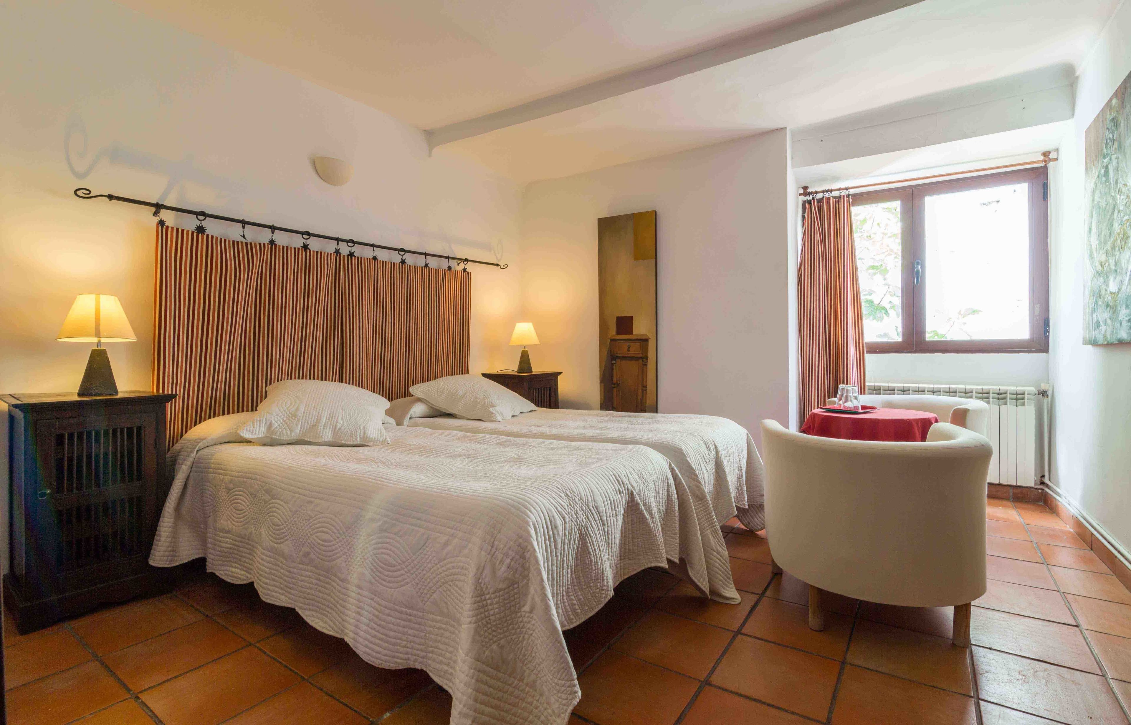 Quarto com duas camas, uma cadeira, uma mesa, radiador e janela com cortinas.