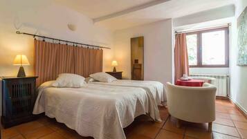 Quarto com duas camas, uma cadeira, uma mesa, radiador e janela com cortinas.