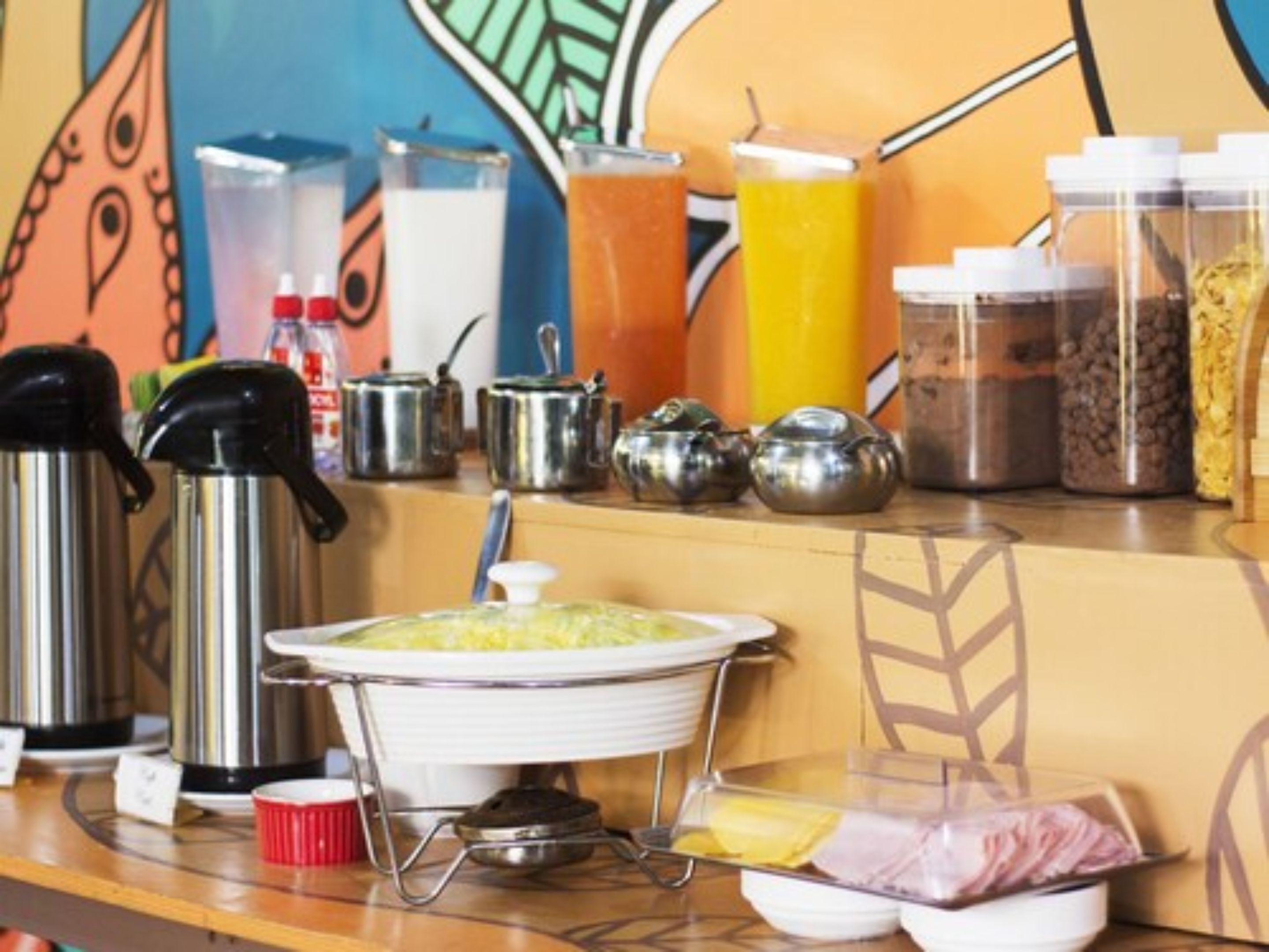 Disposição de buffet com diversos alimentos, incluindo um dispensador de bebida quente, uma panela grande de sopa e recipientes com diferentes tipos de comida.