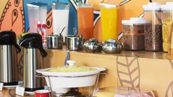 Disposição de buffet com diversos alimentos, incluindo um dispensador de bebida quente, uma panela grande de sopa e recipientes com diferentes tipos de comida.
