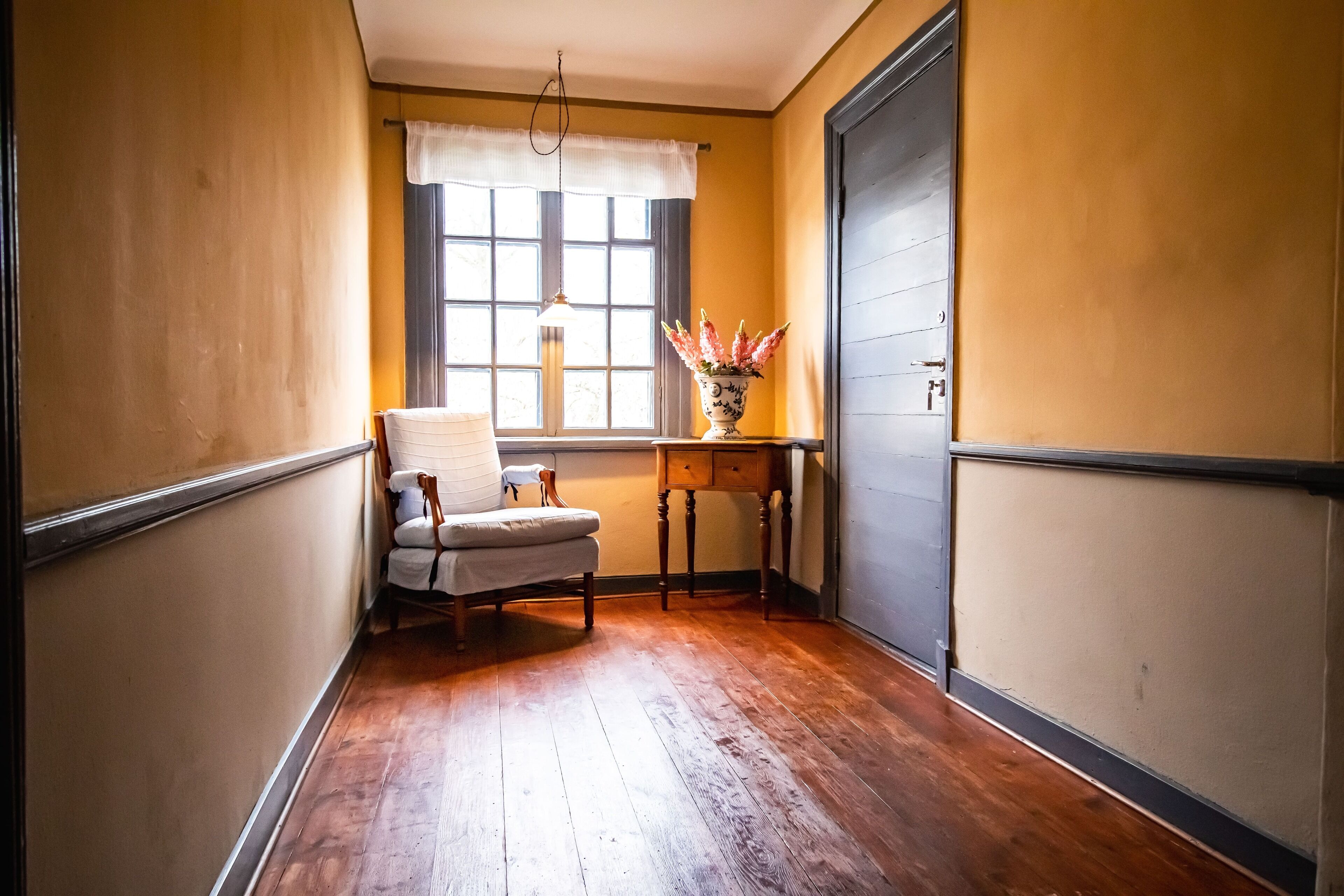 Um corredor estreito com piso de madeira, uma cadeira, uma mesa pequena com um vaso de flores e uma janela com cortina branca.