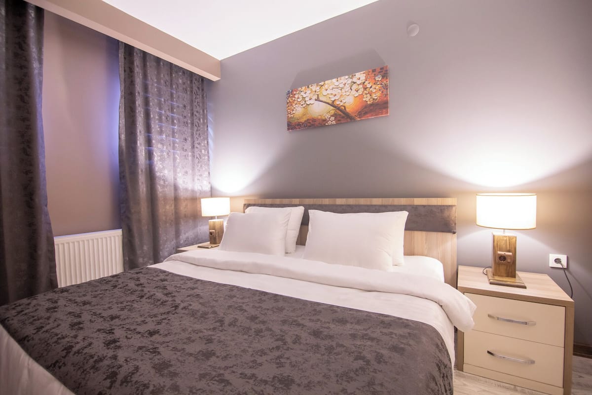 Quarto de hotel com uma cama, duas mesas de cabeceira com luminárias, um radiador e um quadro na parede.