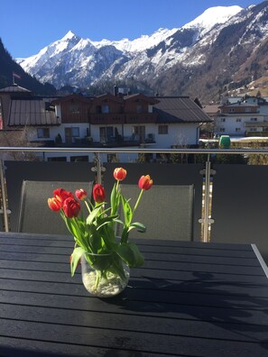 Uma mesa com um vaso de tulipas, uma cadeira e vista para montanhas nevadas e casas ao fundo.