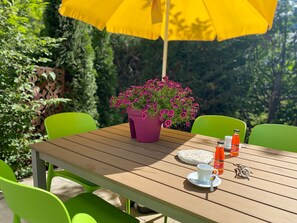 Mesa ao ar livre com guarda-sol amarelo, planta em vaso e louças.