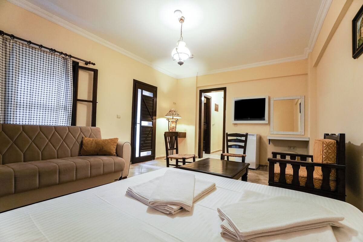 Quarto de hotel com cama, sofá, mesa, cadeira, televisão e janela com cortinas.