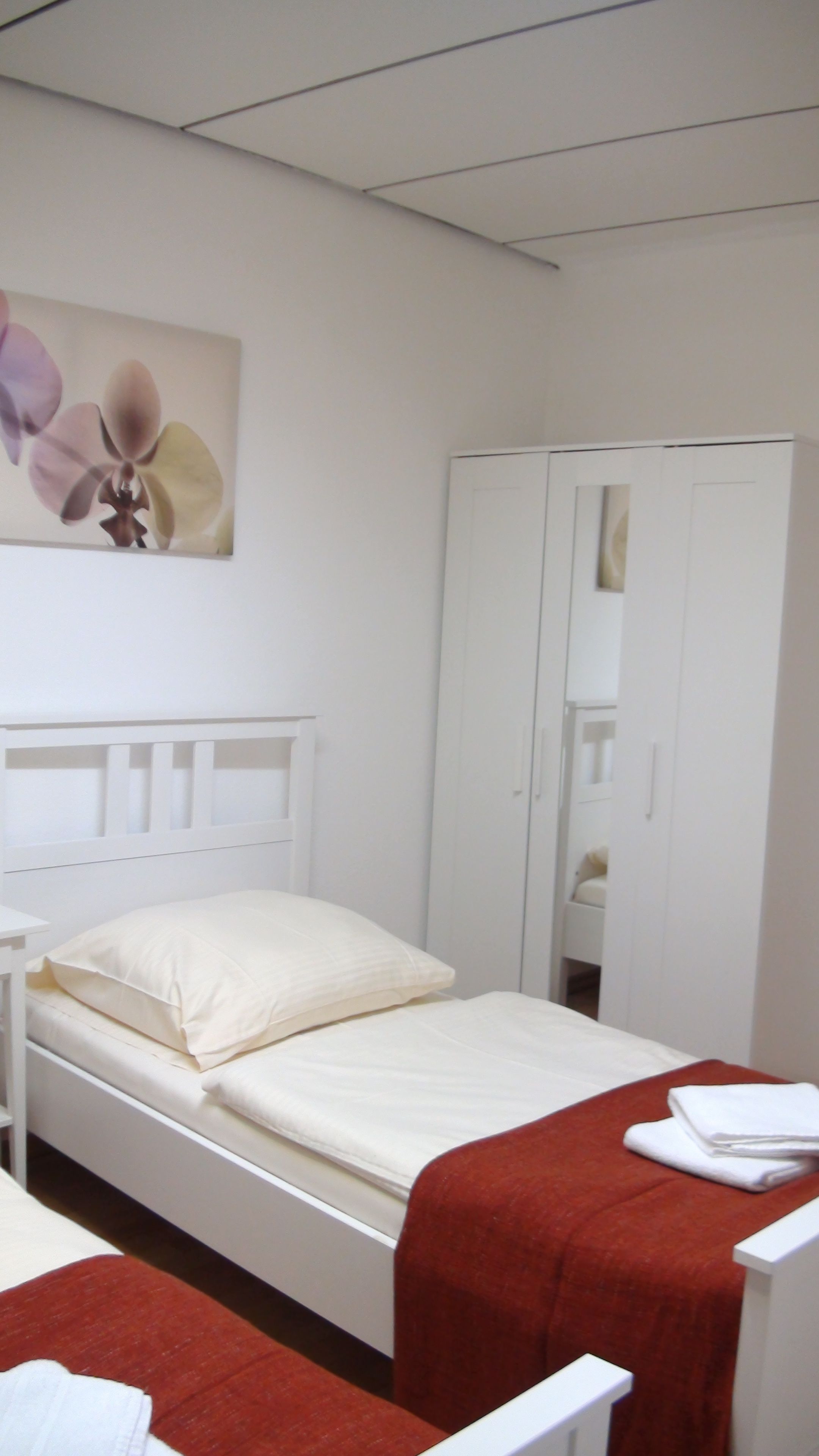 Uma cama de solteiro com cabeceira branca, manta vermelha e travesseiro branco. Há um guarda-roupa branco com espelho e um quadro de flores na parede.