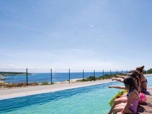Uma piscina com vista para o mar, uma cerca e pessoas sentadas à beira da piscina.