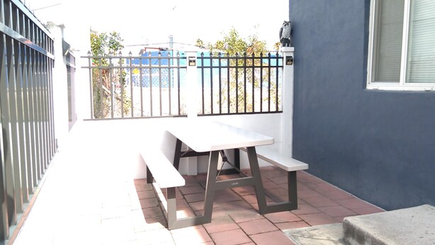 Um pequeno pátio ao ar livre com uma mesa e bancos brancos, uma cerca de metal e um edifício ao fundo.