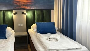Quarto de hotel com duas camas, uma mesinha de cabeceira, cabeceira com iluminação embutida e uma grande pintura de um pôr do sol sobre o oceano.