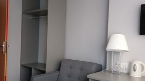 Um quarto com um sofá cinzento, uma mesa com luminária, uma chaleira e um guarda-roupa.