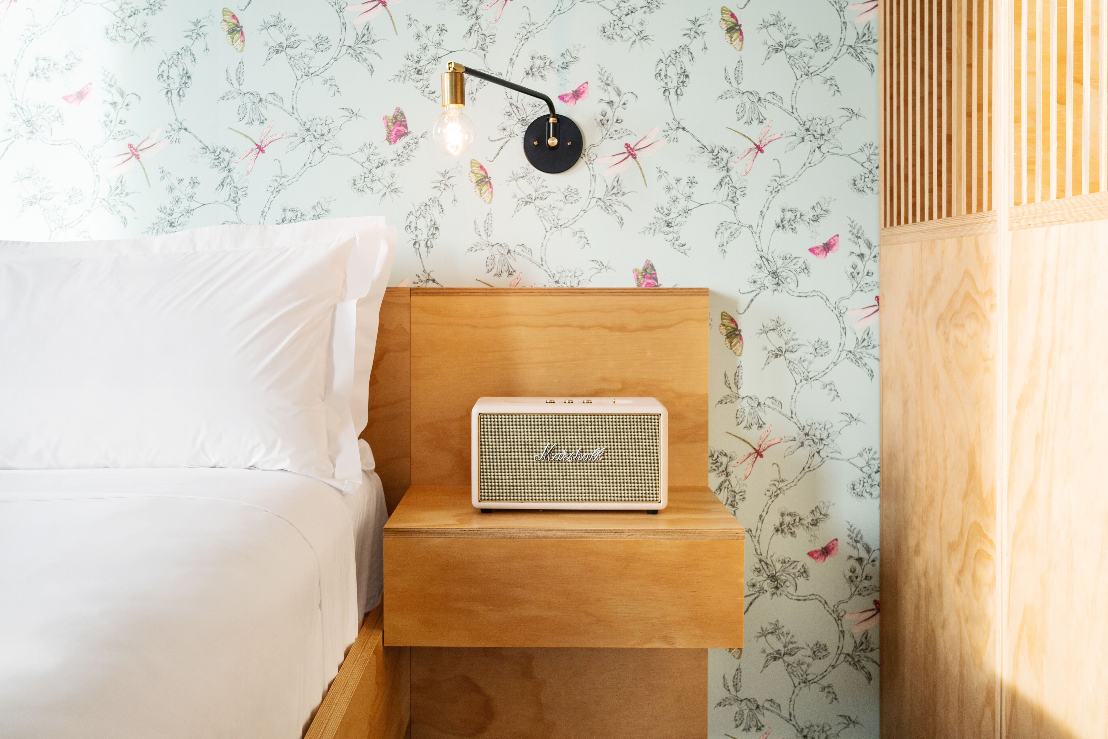 Uma cama com lençóis brancos, uma mesa de cabeceira de madeira com um alto-falante Marshall e uma parede com papel de parede floral.