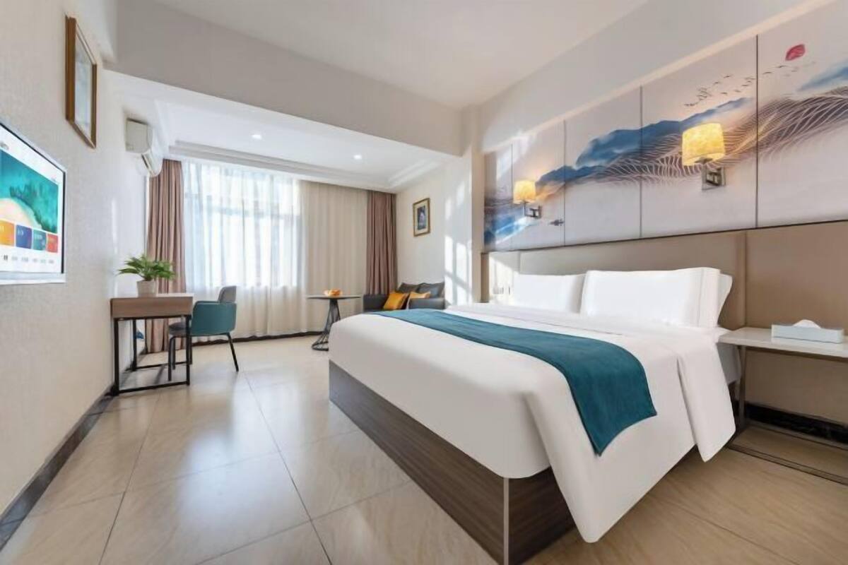 Quarto de hotel com cama, escrivaninha, cadeira, um quadro na parede e janela com cortinas.