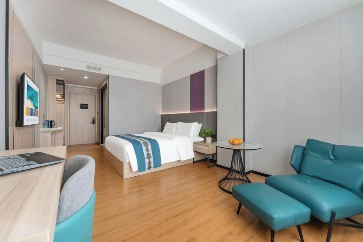 Quarto de hotel moderno com cama, escrivaninha, poltrona, mesinha com uma planta e um sofá.