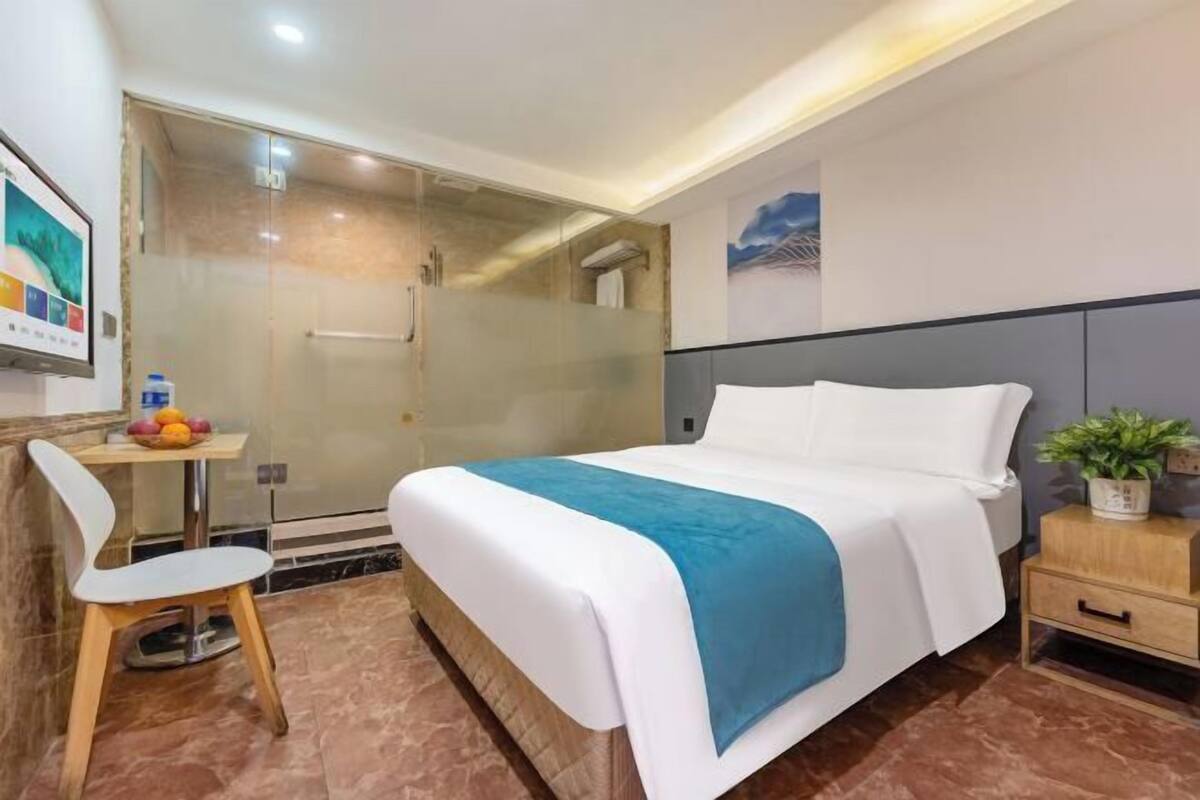 Quarto de hotel com cama, escrivaninha, cadeira, mesa pequena com frutas, planta em vaso e chuveiro.