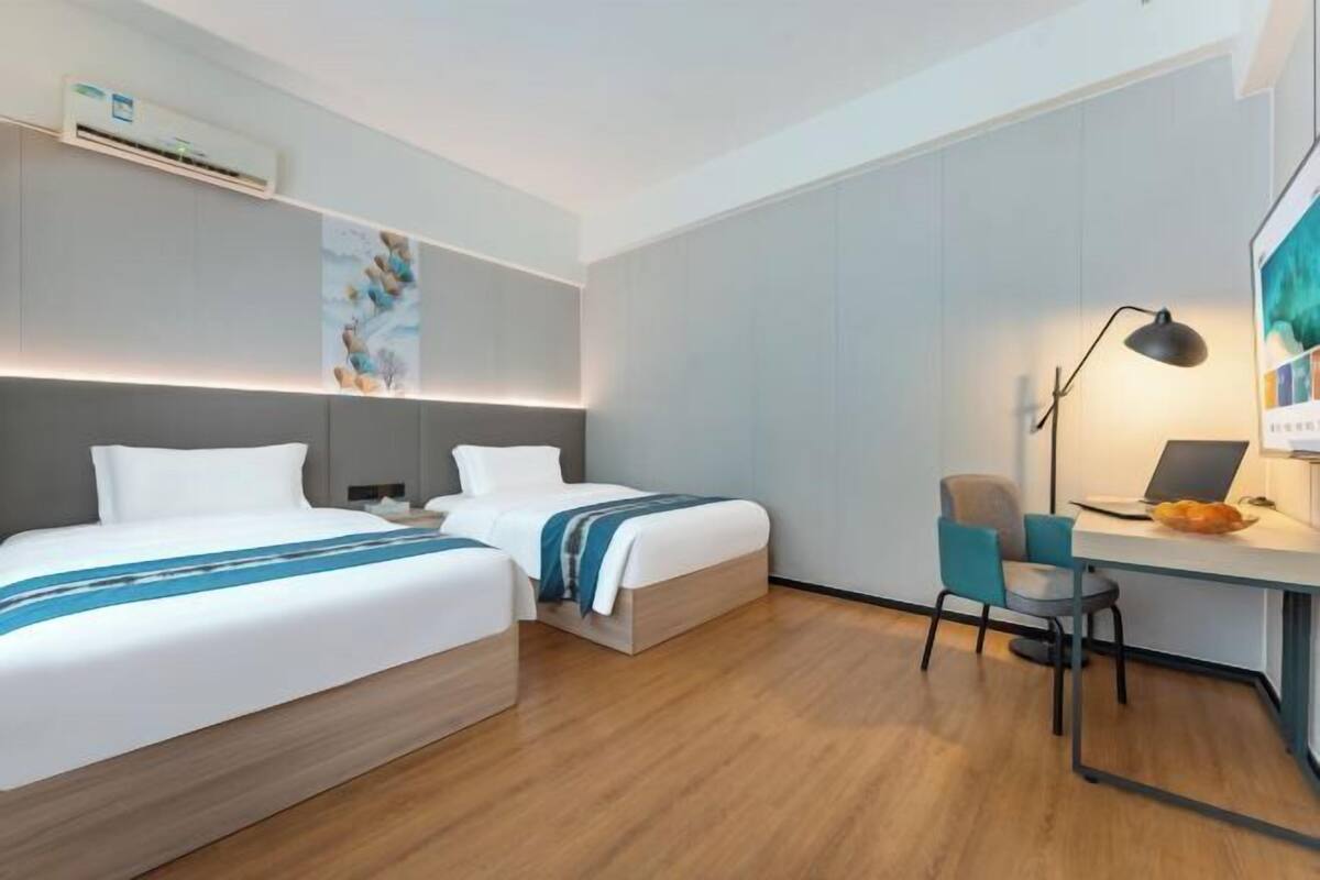 Quarto de hotel moderno com duas camas, uma escrivaninha, uma cadeira e uma luminária de mesa.