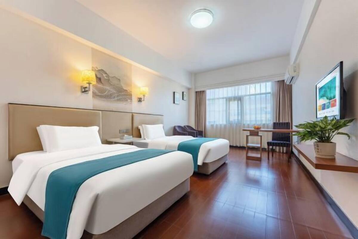 Quarto de hotel com duas camas, uma escrivaninha, uma cadeira, uma televisão e uma planta.