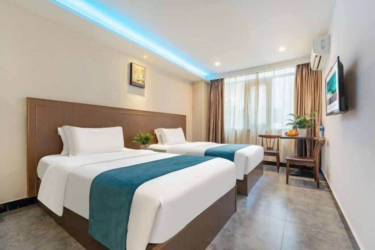 Quarto de hotel com duas camas, uma mesinha com uma cadeira, televisão e janela com cortinas.