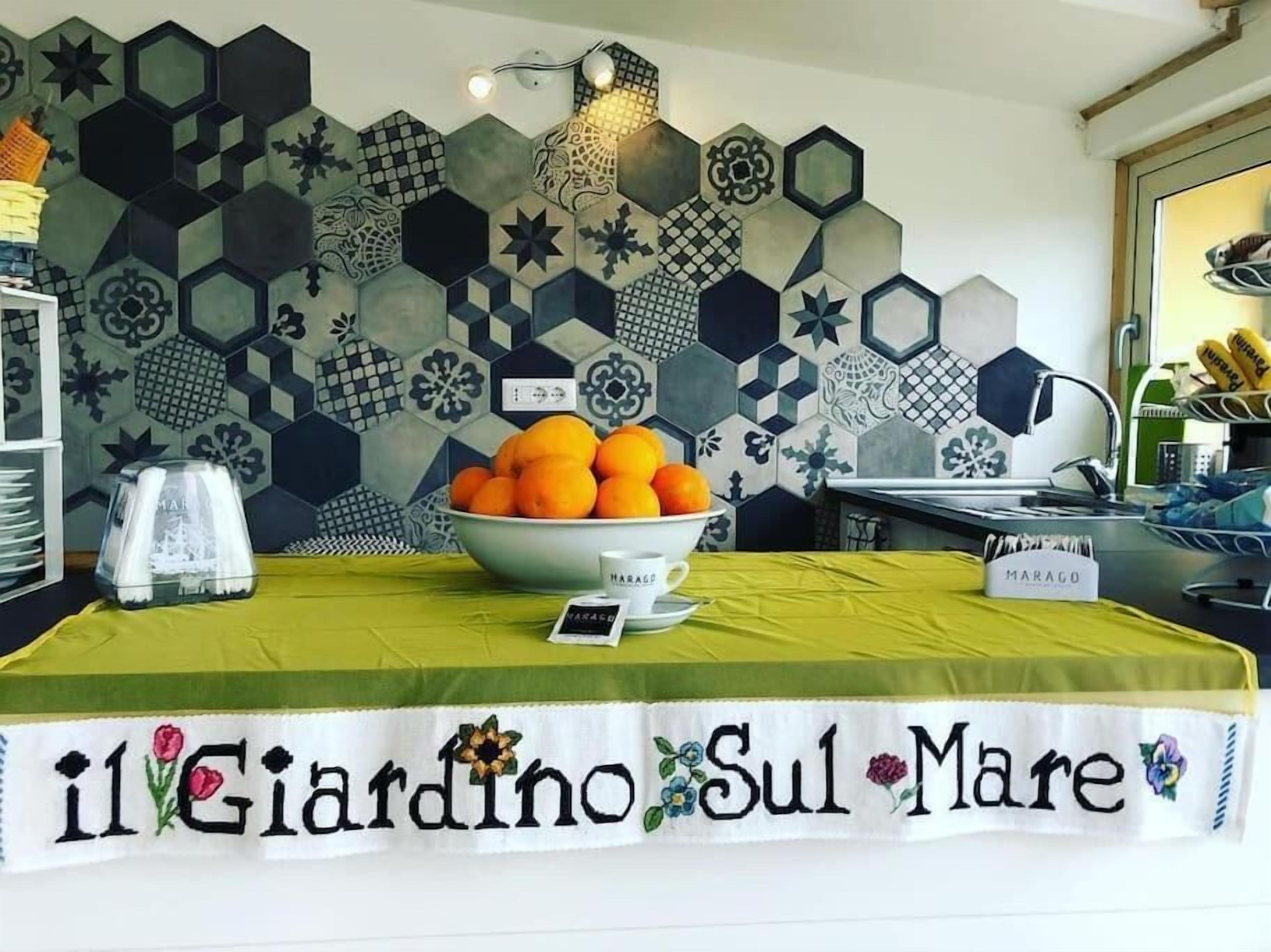 Uma mesa com toalha verde, uma tigela de laranjas e uma placa com o texto 'il Giardino Sul Mare'.