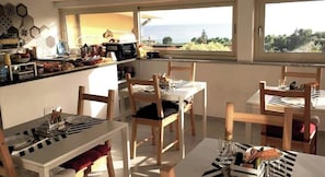 Espaço de refeições com duas mesas e cadeiras, cozinha equipada com diversos eletrodomésticos e utensílios, e janelas amplas com vista para o verde.