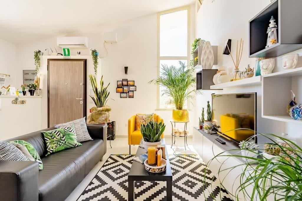 Uma sala de estar moderna com sofá, mesa de centro e televisão. Há plantas, uma porta e uma estante com objetos decorativos.