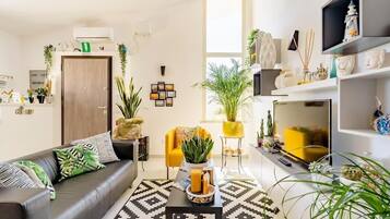 Uma sala de estar moderna com sofá, mesa de centro e televisão. Há plantas, uma porta e uma estante com objetos decorativos.