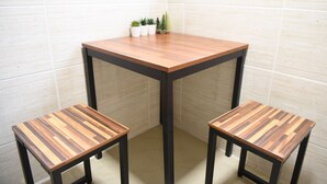 Uma pequena mesa quadrada com tampo de madeira e pernas de metal preto, acompanhada por dois taburetes combinando.