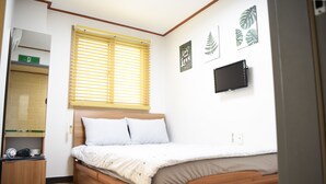 Uma cama bem arrumada com estrutura de madeira e roupa de cama branca, uma mesa de cabeceira com abajur e uma janela com persianas.