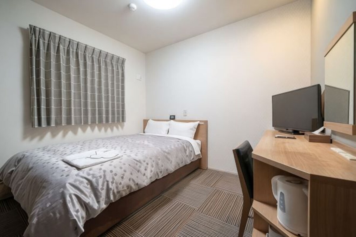 Quarto com cama, escrivaninha, cadeira, televisão e chaleira.