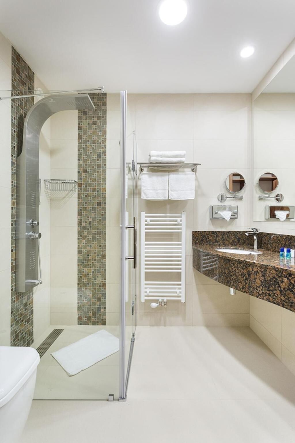 Uma casa de banho moderna com cabine de duche em vidro, um porta-toalhas branco, lavatório com bancada de granito e um espelho grande.