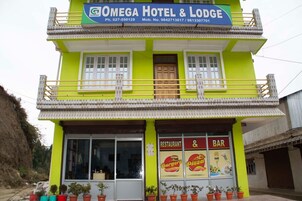 Um edifício de dois andares com uma placa para Omega Hotel & Lodge. Possui restaurante e bar no andar térreo, com plantas em vasos na frente.