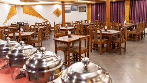 Um restaurante com mesas e cadeiras de madeira, toalha de mesa vermelha e travessas de servir prateadas.