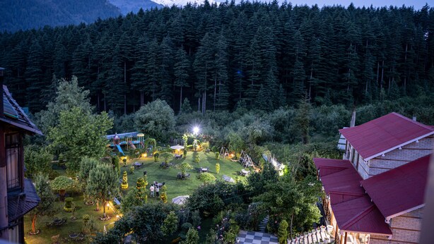 Um jardim bem cuidado, com diversas árvores e arbustos, uma pequena área de recreação infantil e um espaço para refeições.