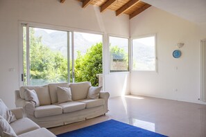 Uma sala de estar espaçosa com um sofá branco grande, um tapete azul e grandes janelas com vista para as montanhas.