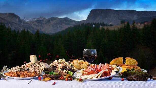 Uma mesa com diversos alimentos e um copo de vinho tinto, com montanhas ao fundo.