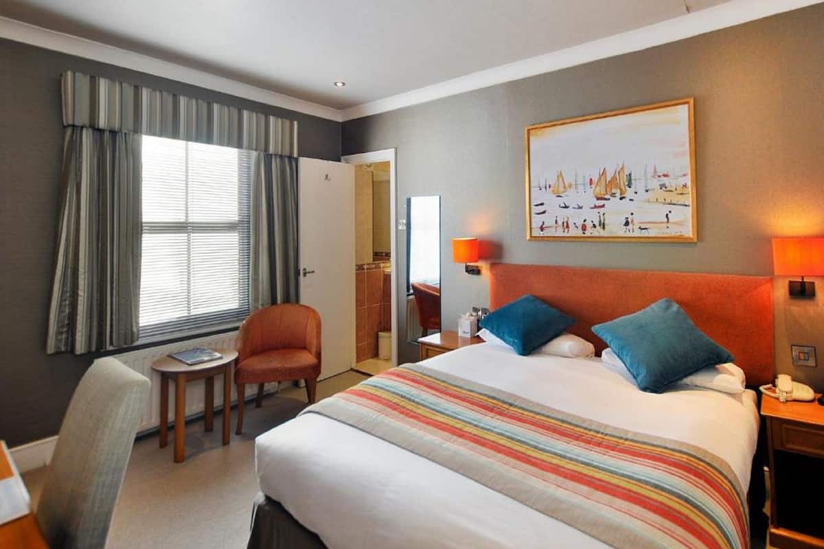 Quarto de hotel com cama, escrivaninha, cadeira, janela com cortinas, quadro na parede e mesa de cabeceira com abajur.