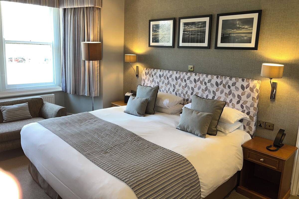 Quarto de hotel com uma cama grande, duas mesas de cabeceira, uma janela com cortinas e três quadros na parede.