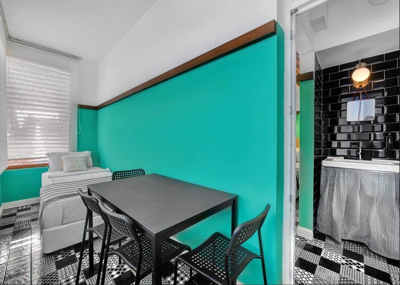 Quarto compacto com uma parede em tom de verde-água, uma pequena mesa de jantar com quatro cadeiras, cama com roupa de cama branca e chão quadriculado em preto e branco.
