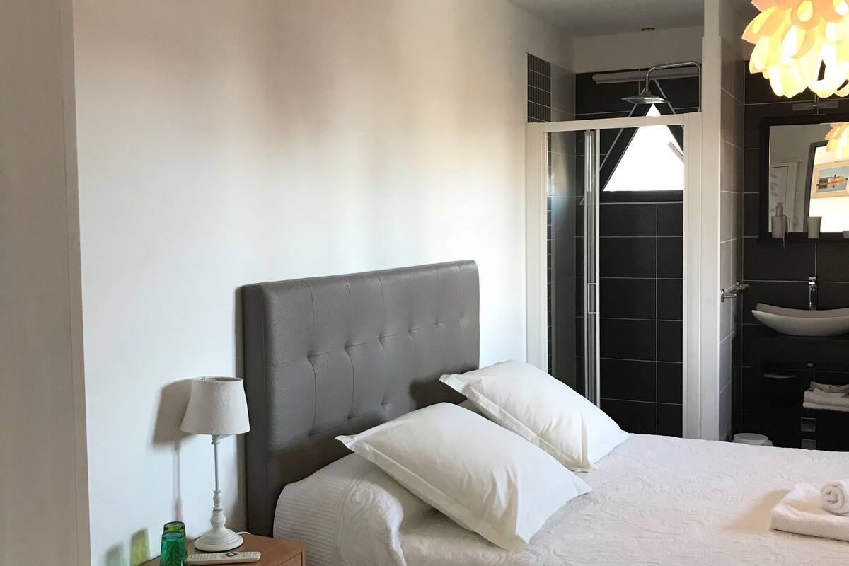 Quarto com cama, mesa de cabeceira, luminária, mesa de cabeceira de madeira, espelho, pia e chuveiro.