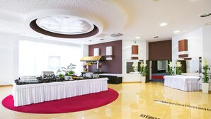 Um amplo saguão de hotel com uma recepção central, espaço para buffet e um tapete vermelho que leva à entrada.