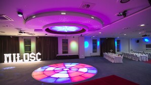 Uma sala de conferências com uma instalação de iluminação circular, um palco com a sinalização 'MILOSC' e fileiras de mesas com toalhas brancas.